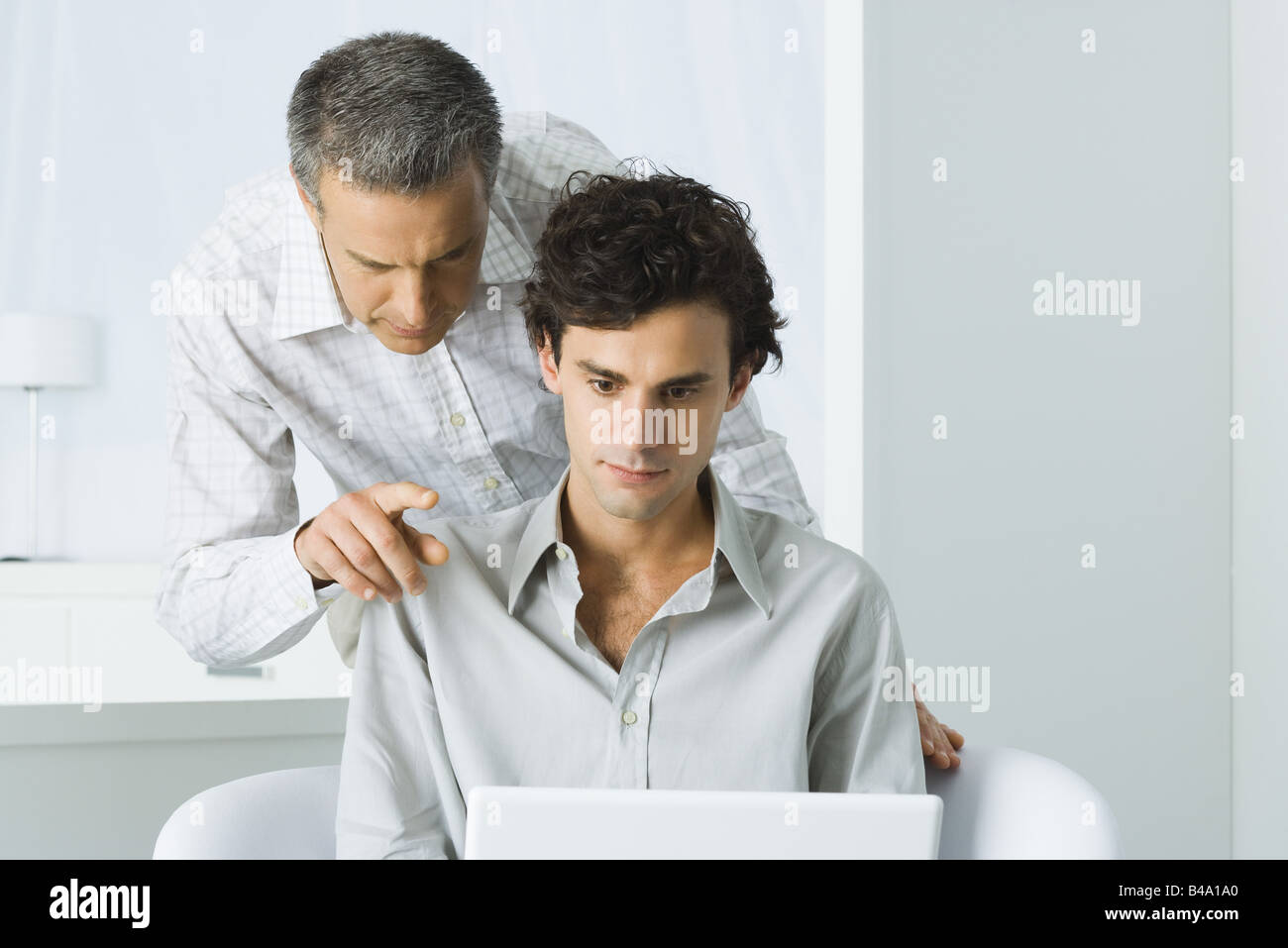 Mature man leaning over young man's shoulder, both looking at laptop ...