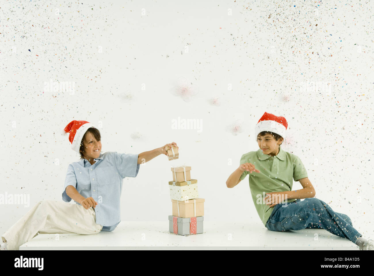 Two boys wearing Santa hats stacking presents, confetti falling around ...