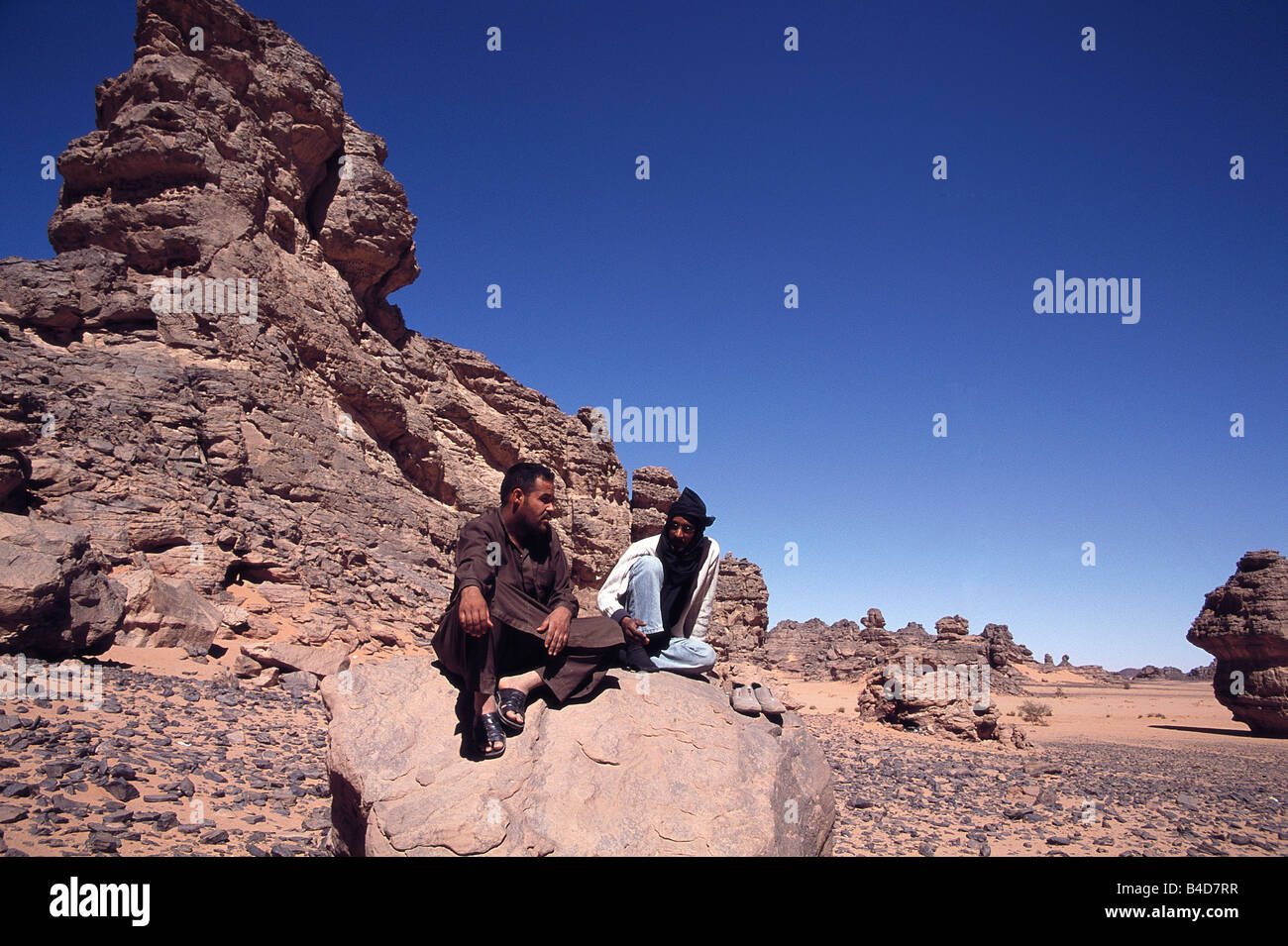 Two Libyans sit in the sun on a rock and chat in the Sahara Desert ...