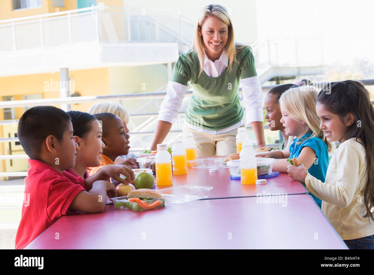 Teacher leaning on table outdoors while students eat lunch (high key ...