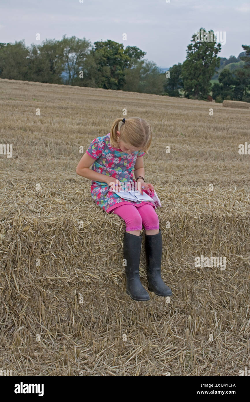 Young girl sat on a straw bale in stubble field reading and drawing ...
