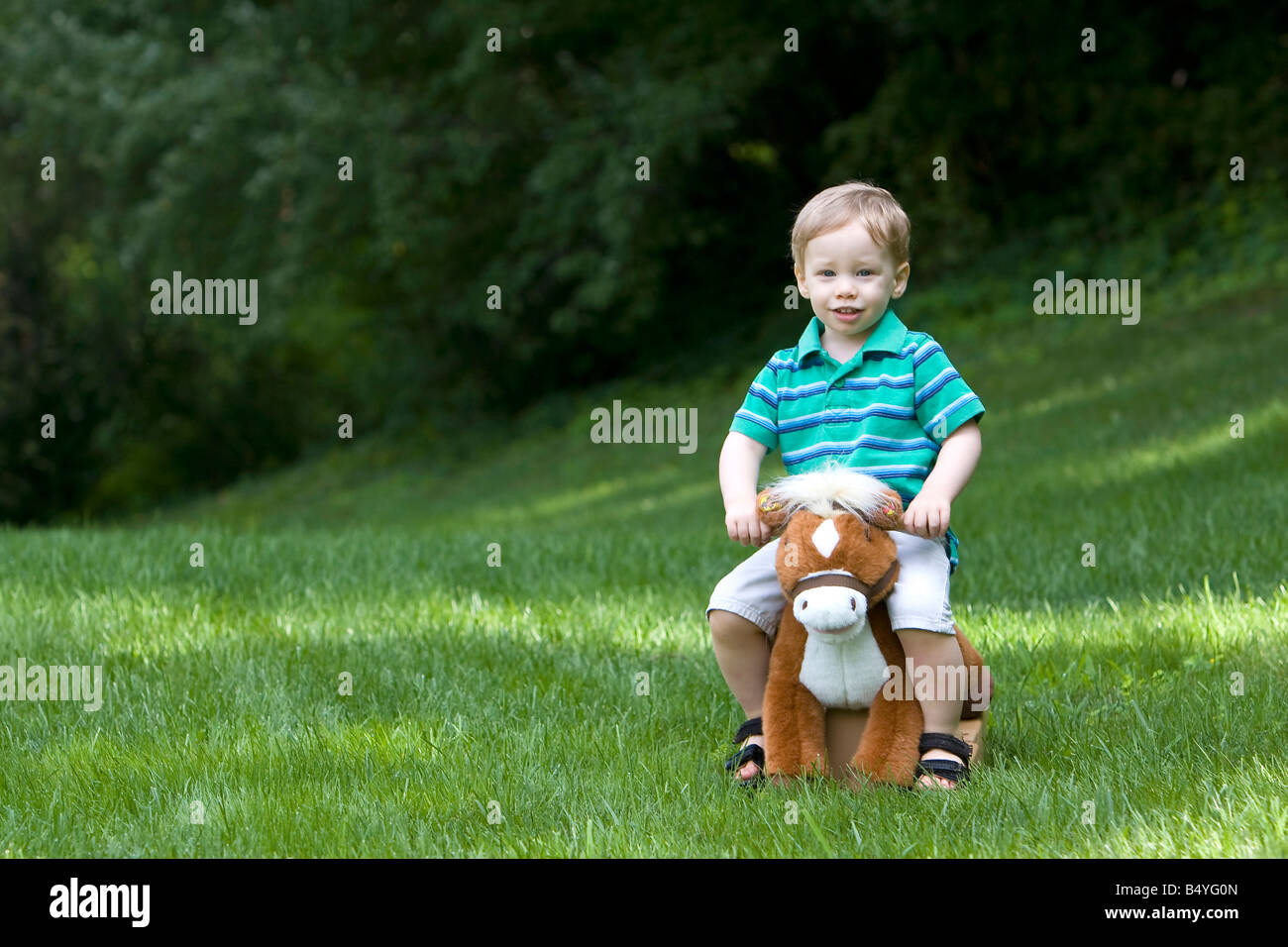 Boy riding a toy rocking horse in a field with trees in background ...