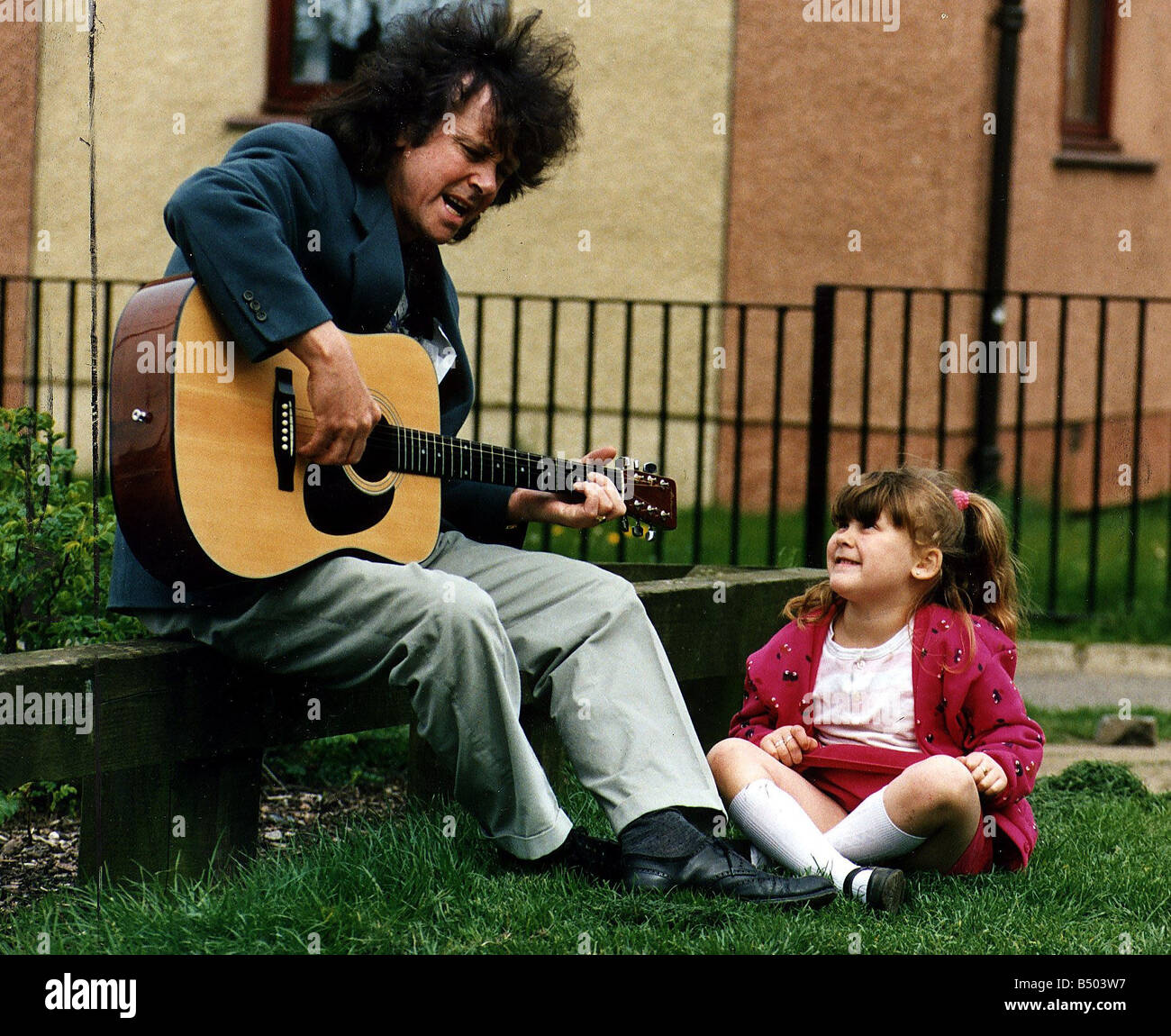 Donovan playing guitar and singing on low wall watched by small girl ...