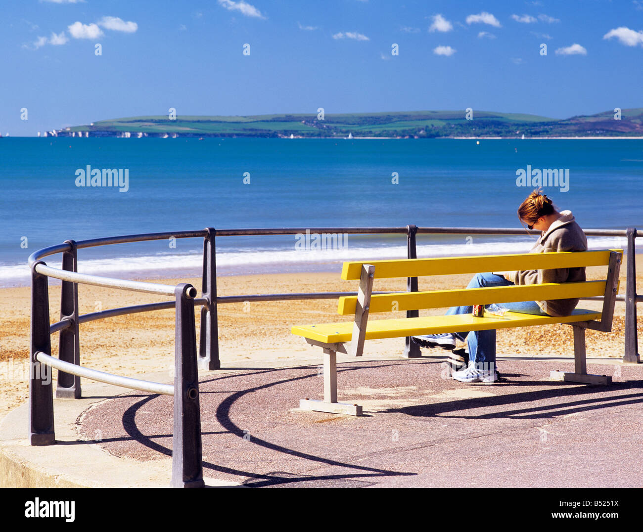 Young woman sat quietly reading on yellow seafront bench overlooking a ...
