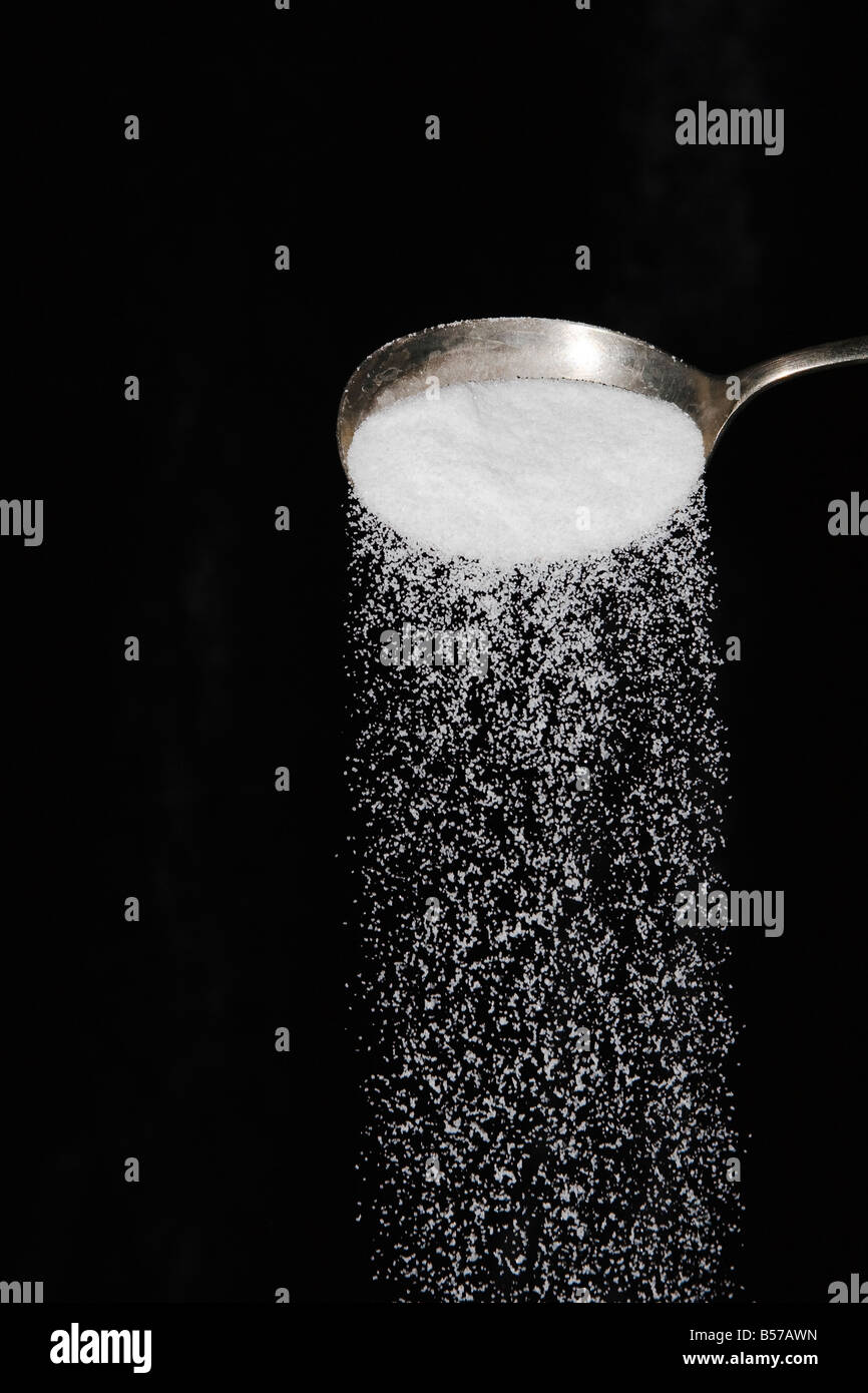 Sugar falling from a silver coloured spoon against a black background ...