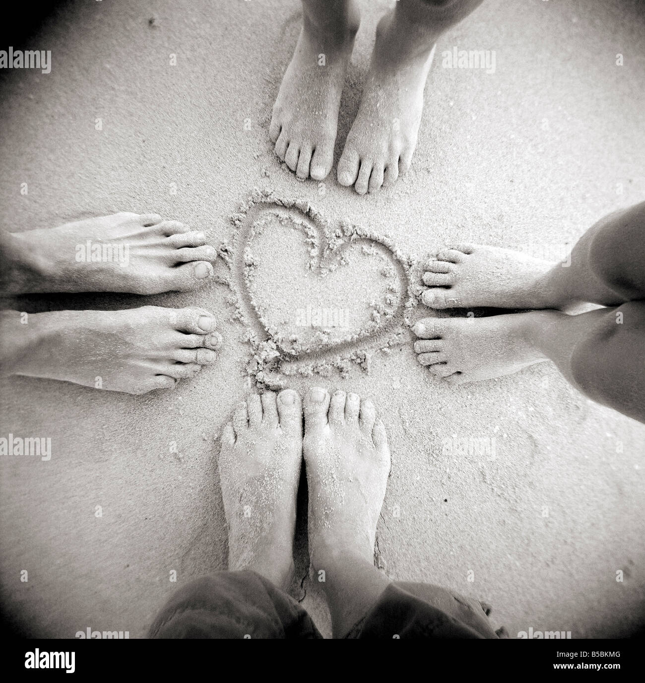 Four pairs of feet standing around a heart shape drawn in sandy beach ...