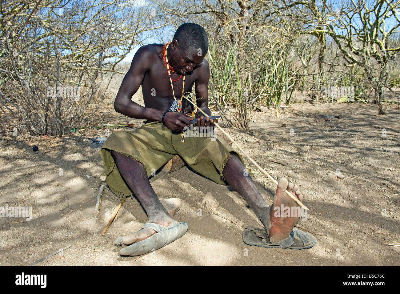 A member of the Hadza tribe shapes a hunting arrow with a knife Lake ...
