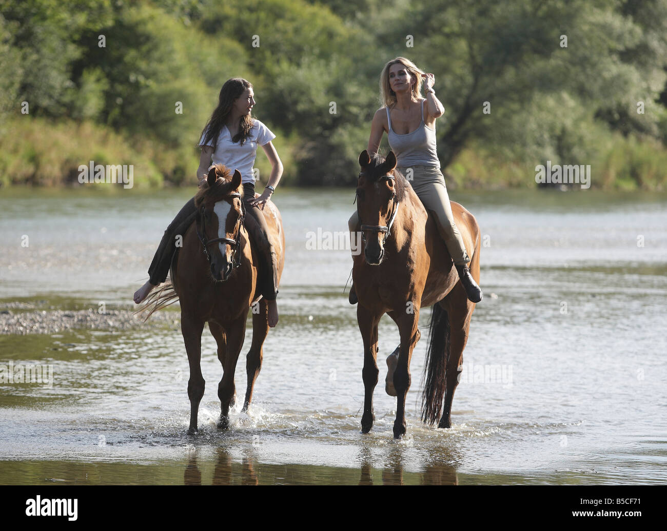 Blonde woman and teenage girl riding horseback in a shallow riverbed ...