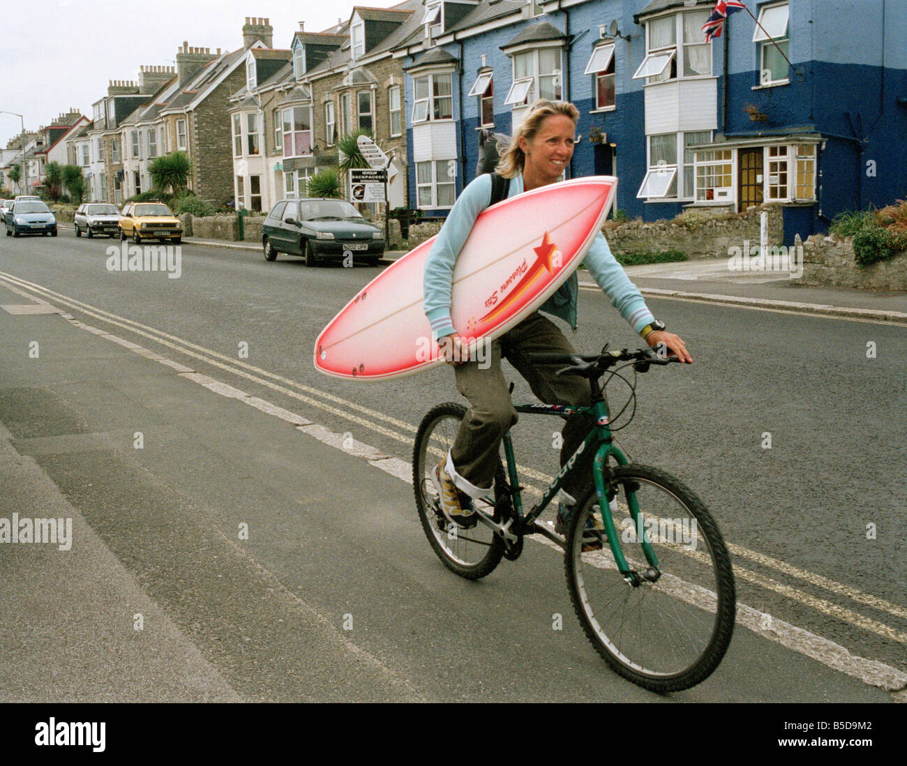 Female surfer cycling to Fistral Beach holding her pink short board ...