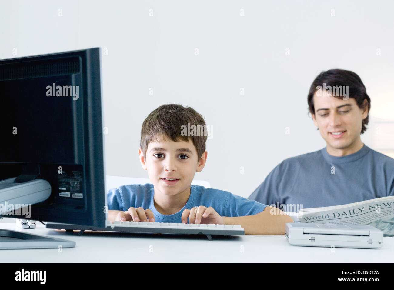 Little boy using computer, smiling at camera, father reading newspaper ...