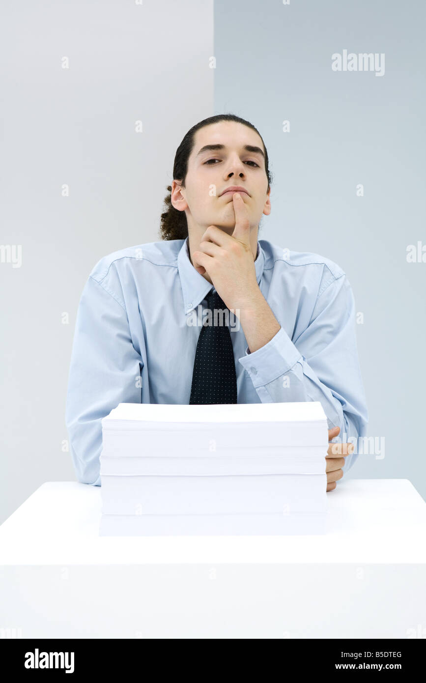Young office worker behind stack of papers, hand under chin, looking at ...