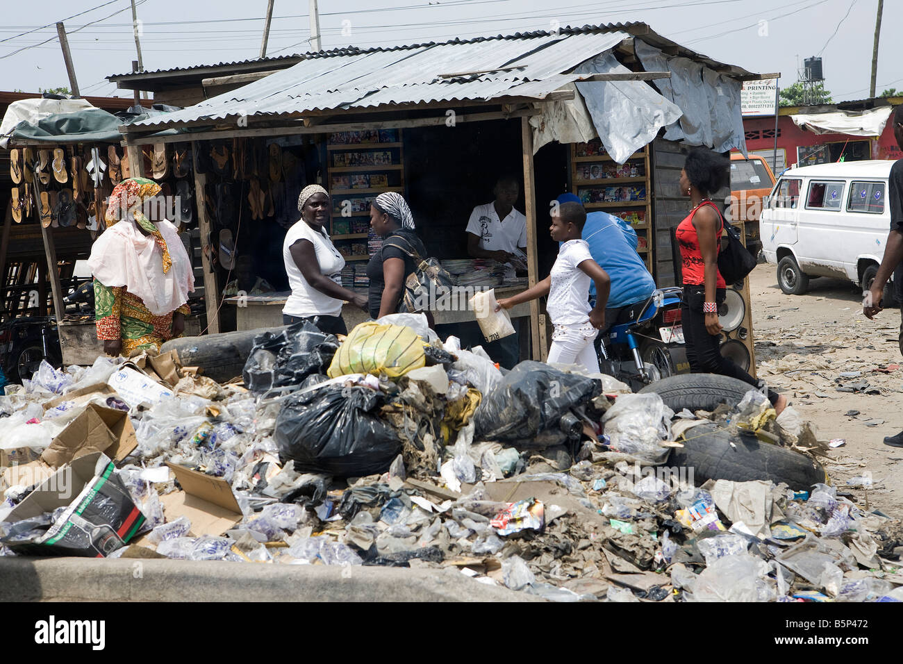 A crowd of people walk past a Nigerian market stall which has a rubbish ...