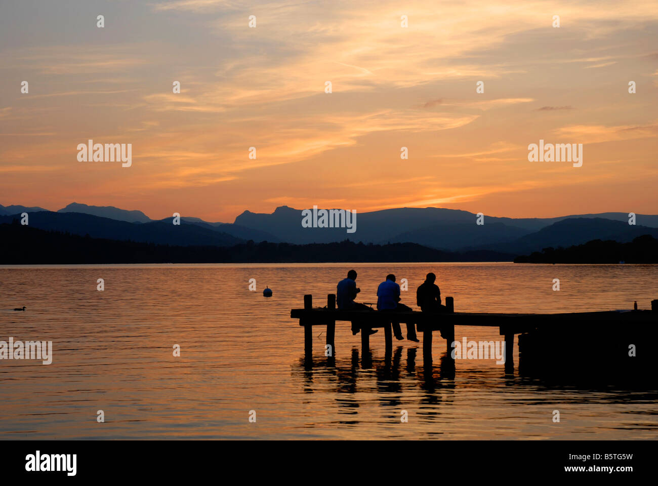 Three boys sitting on a jetty and fishing at sunset, Lake Windermere ...