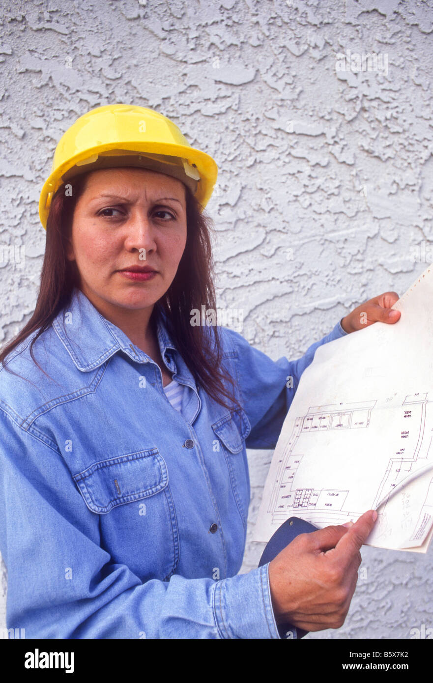 Hispanic woman engineer wearing hard hat examines blueprints of project ...