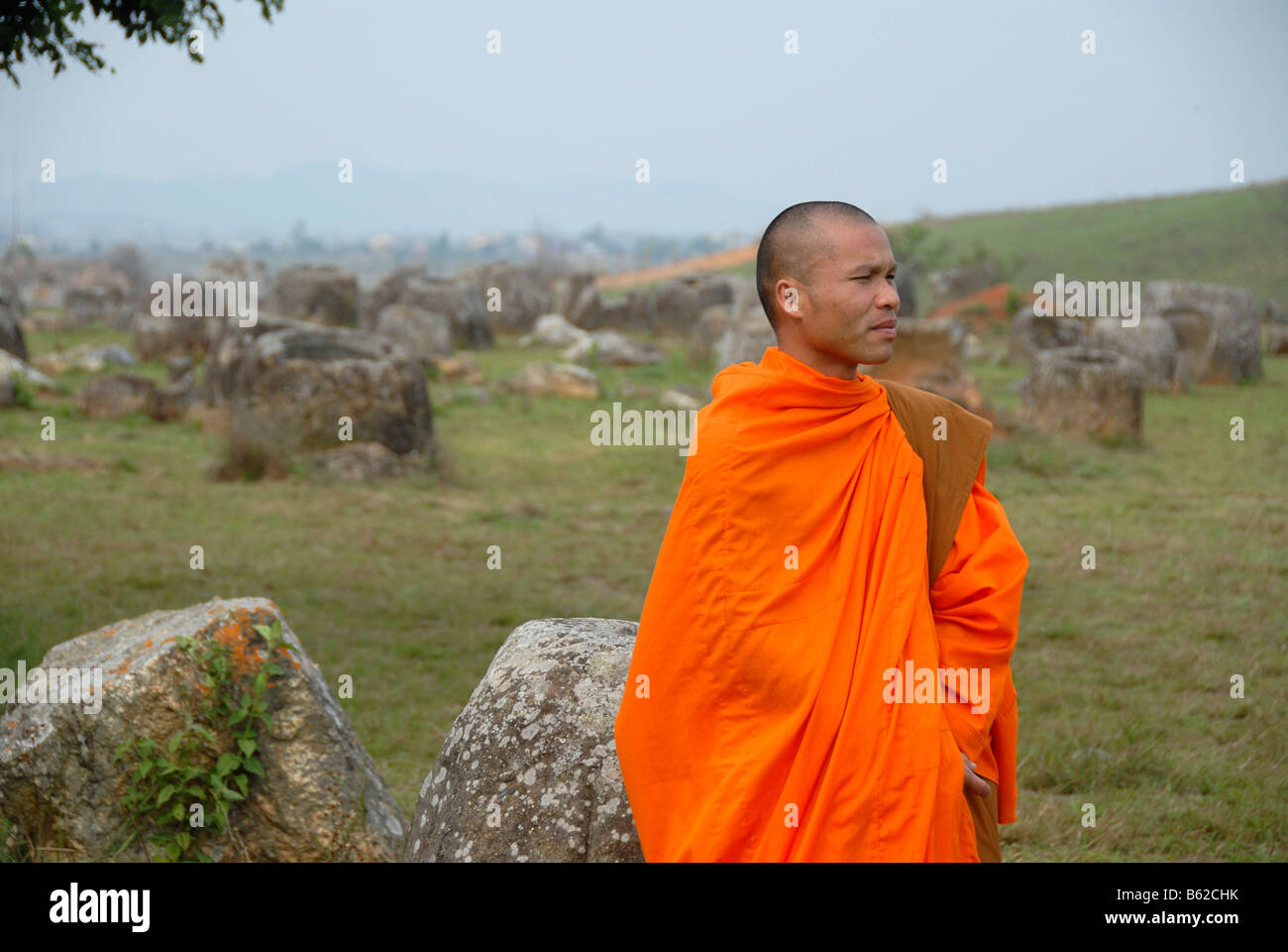 Young monk wearing an orange cowl standing in front of enormous jars ...