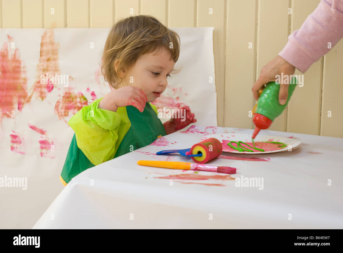 A two year old girl in a plastic smock painting in a playroom with a ...