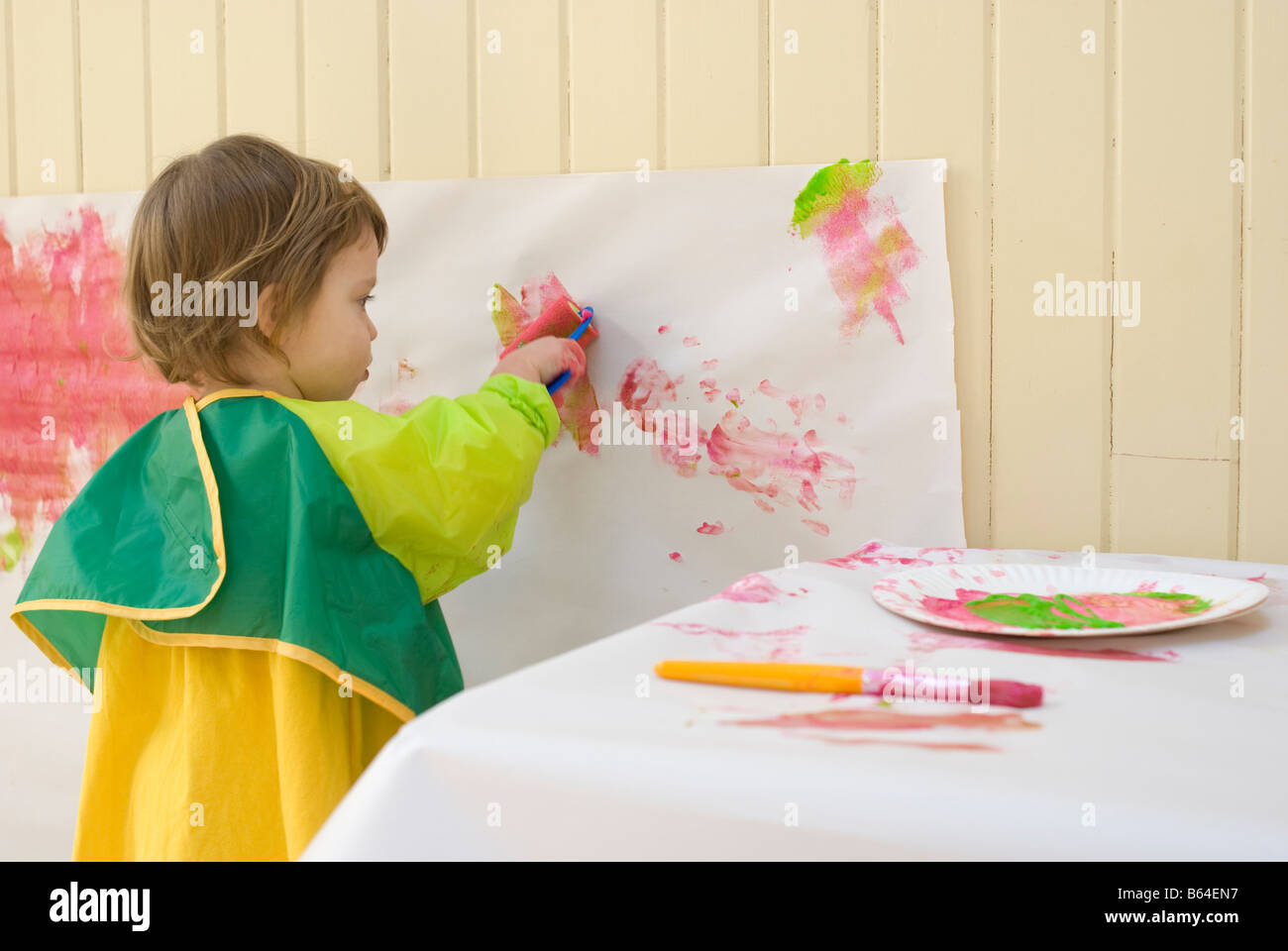 A two year old girl in a plastic smock painting in a playroom with a ...
