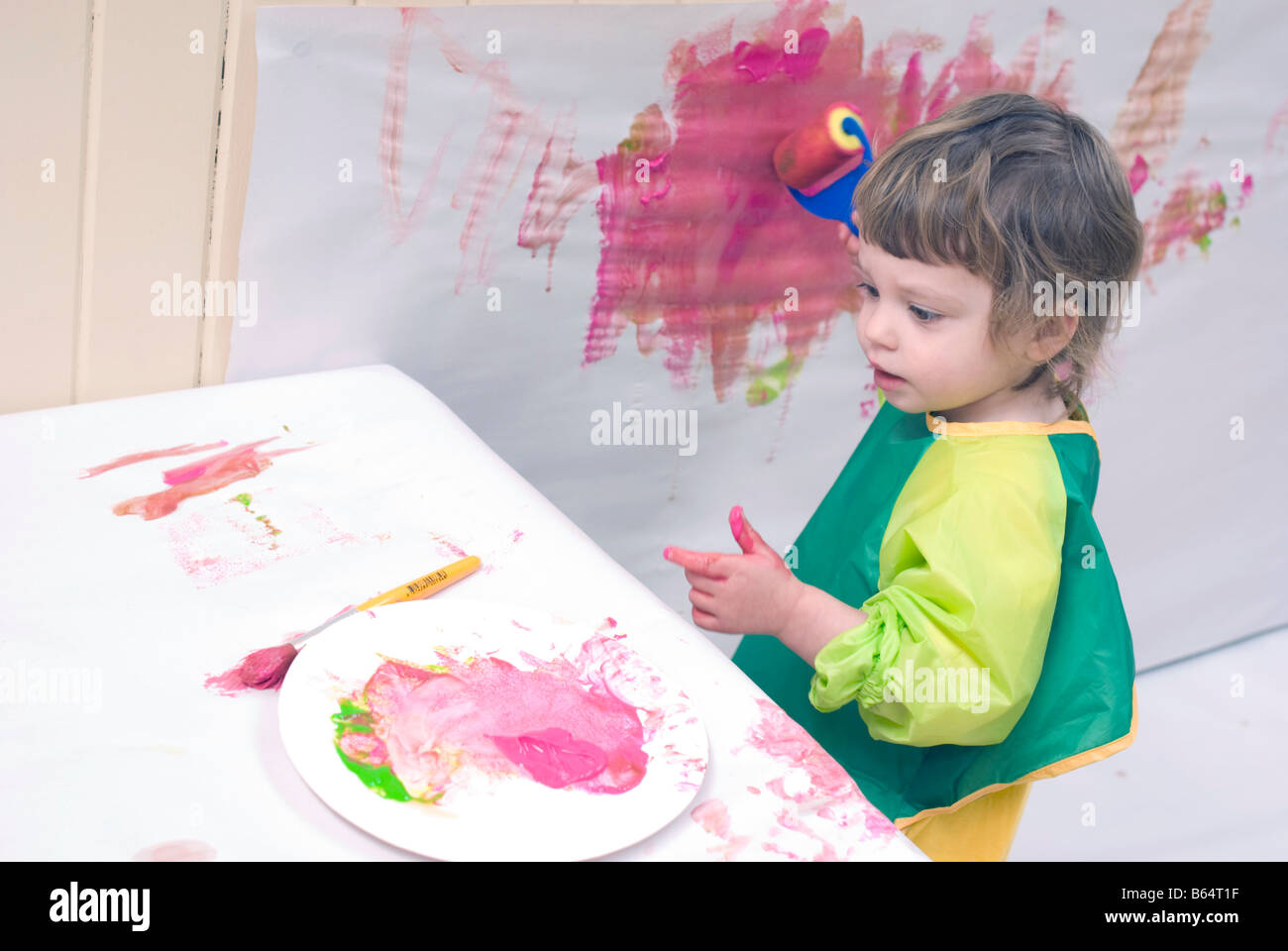 A two year old girl in a plastic smock painting in a playroom with a ...
