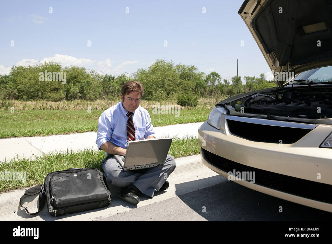 A businessman sitting on the curb working beside his broken down car ...