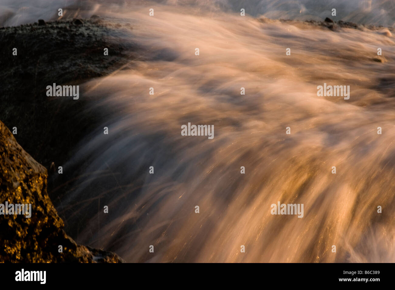 An incoming wave washing over rocks, lit by the setting sun. Taken at ...
