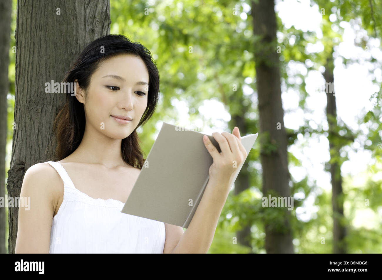 Young woman leaning against tree reading book Stock Photo - Alamy