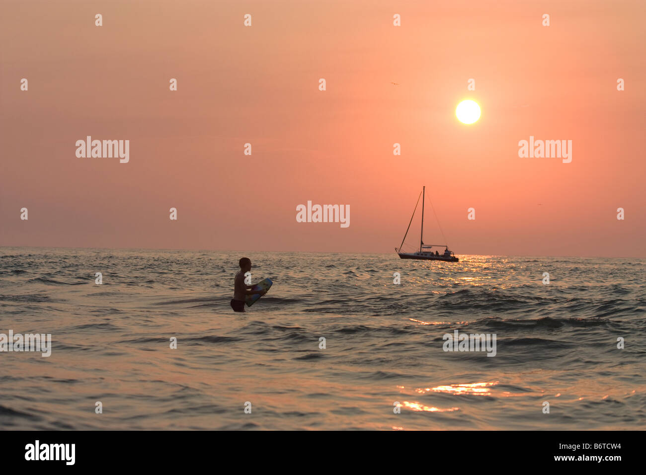 Boy swimming in shallow water at sunset with sailboat in background ...