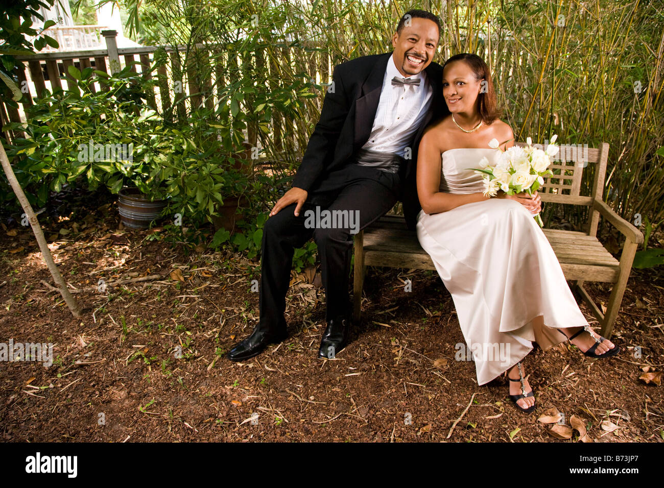 Happy African-American bride and groom on bench outdoors on wedding day ...