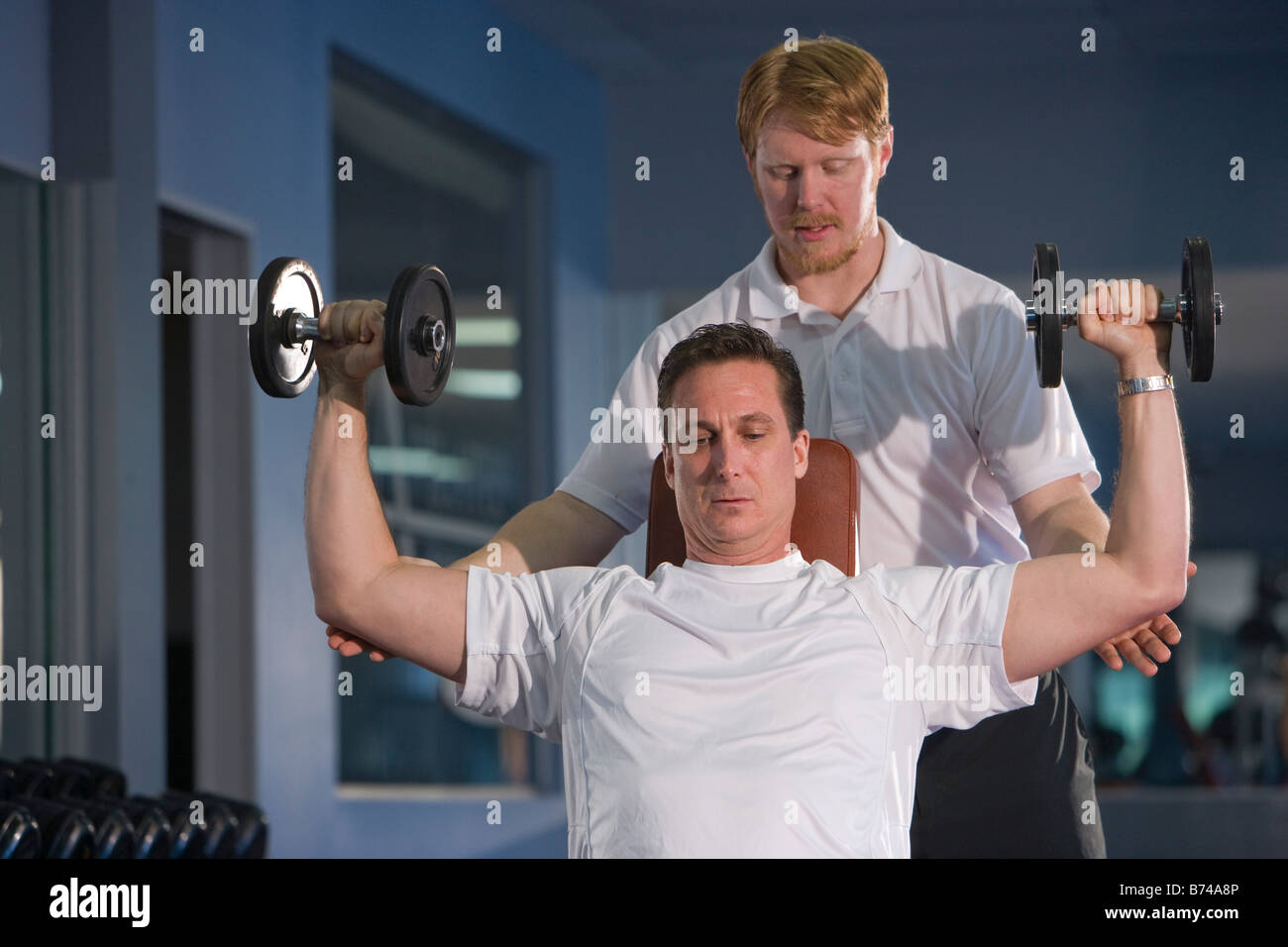 Middle-aged man lifting weights at fitness center with personal trainer ...