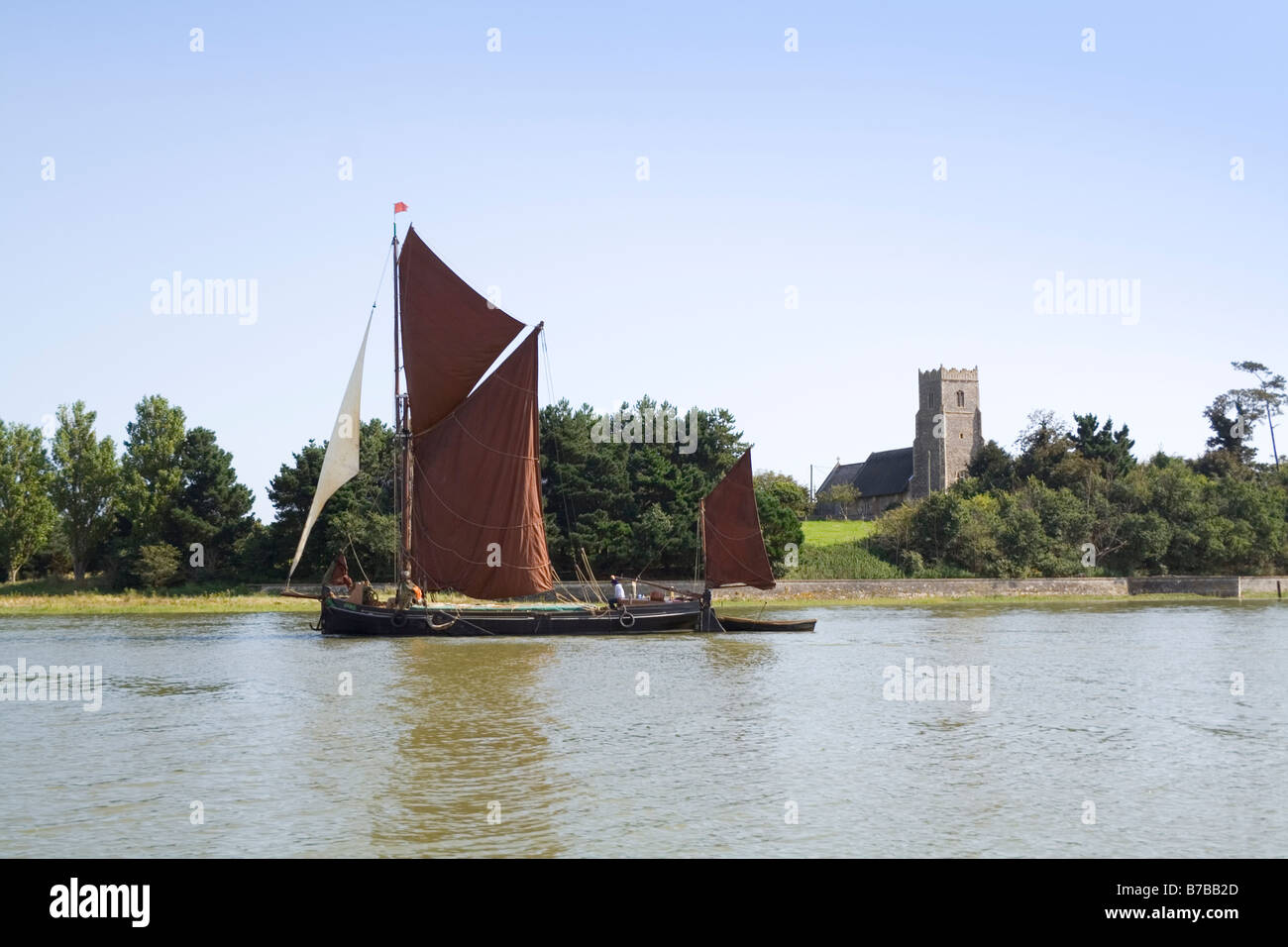 A Victorian spritsail barge glides under full sail past Iken Church on ...