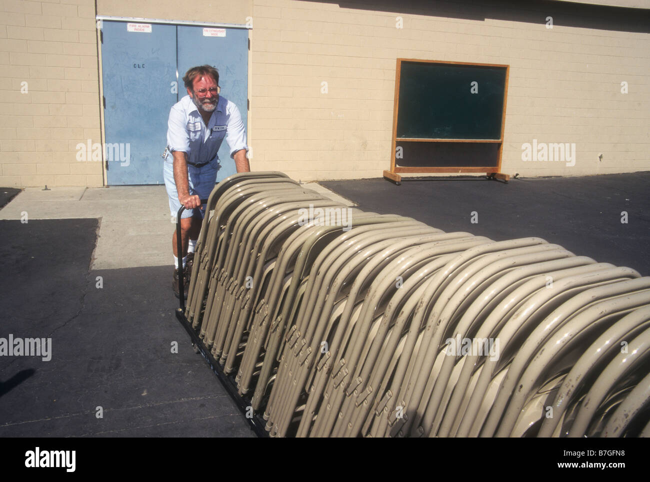 Male custodian pushes rack of folding metal chairs across campus of an ...