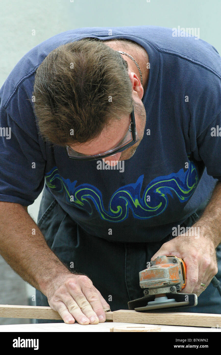 A man wearing safety goggles and sanding wood with an electric sander ...