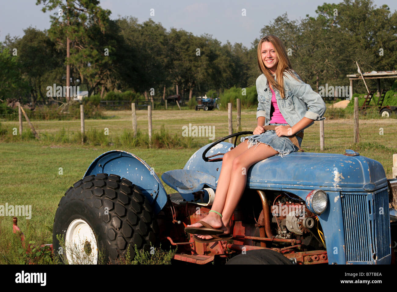 A beautiful blond girl sitting on a tractor in the middle of a farm ...