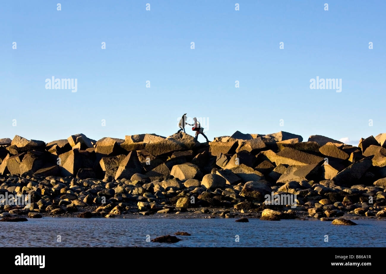 Two people crossing a rock jetty made of large boulders at low tide ...