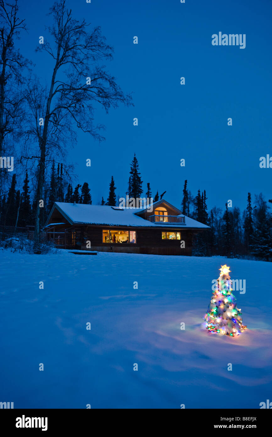 Lit Christmas tree in snow outside a log home during winter at twilight ...