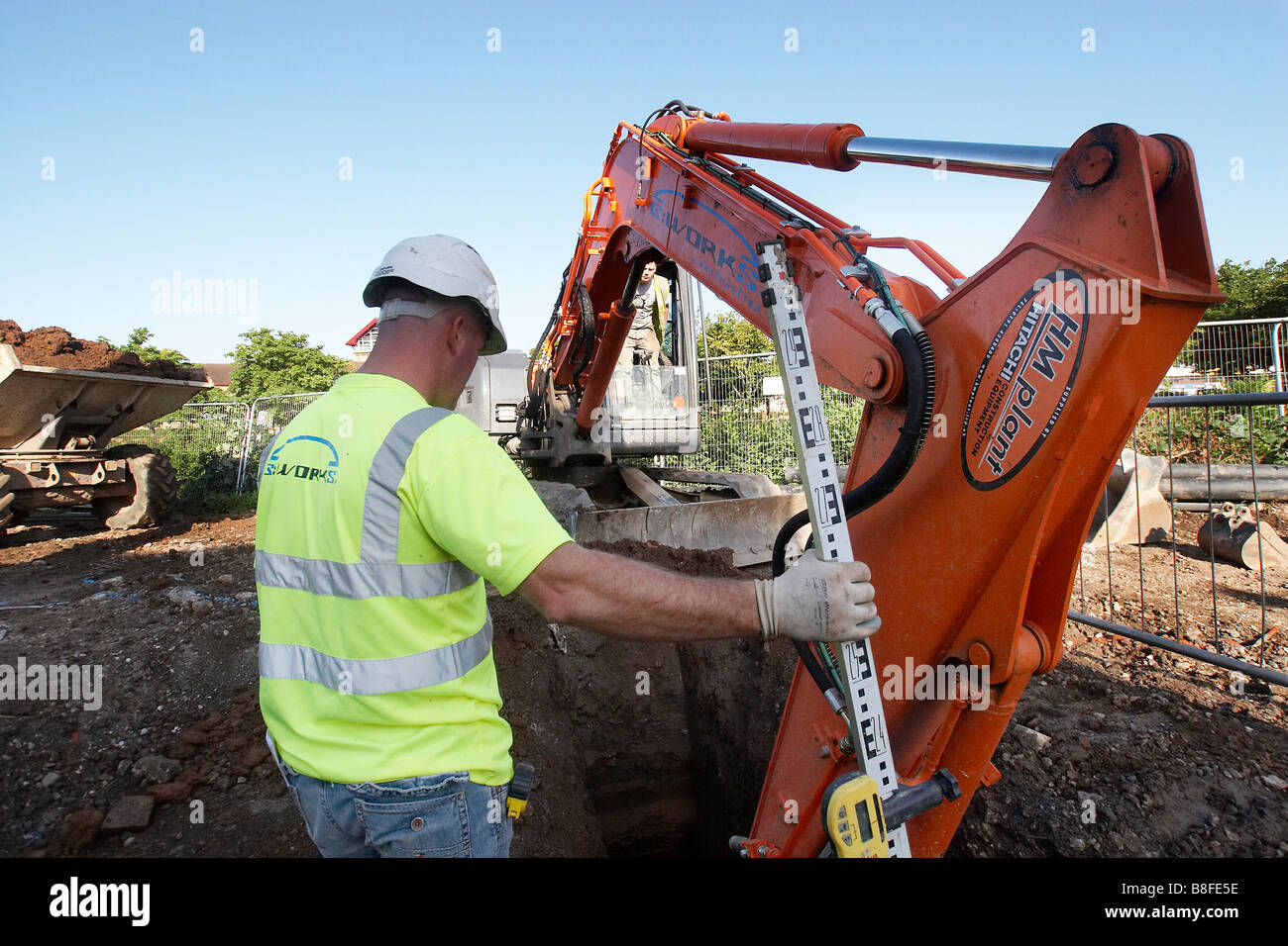Jcb digging a trench on a construction site while a workman measures ...