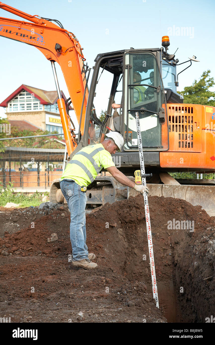 Jcb digging a trench on a construction site while a workman measures ...