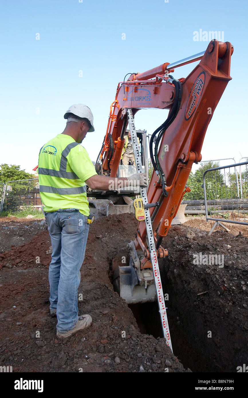 Jcb digging a trench on a construction site while a workman measures ...
