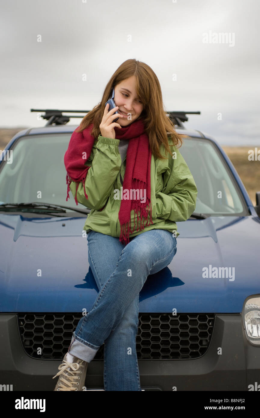 Teen girl sitting on the hood of a car calling or texting on her cell ...