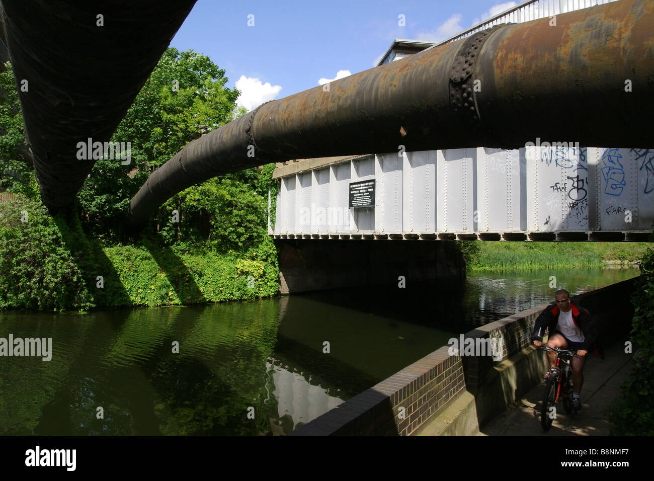 A cyclist on a canal path below a bridge and industrial pipe Stock ...