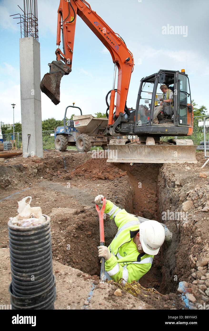 Jcb digging a trench on a construction site while a workman digs Stock ...