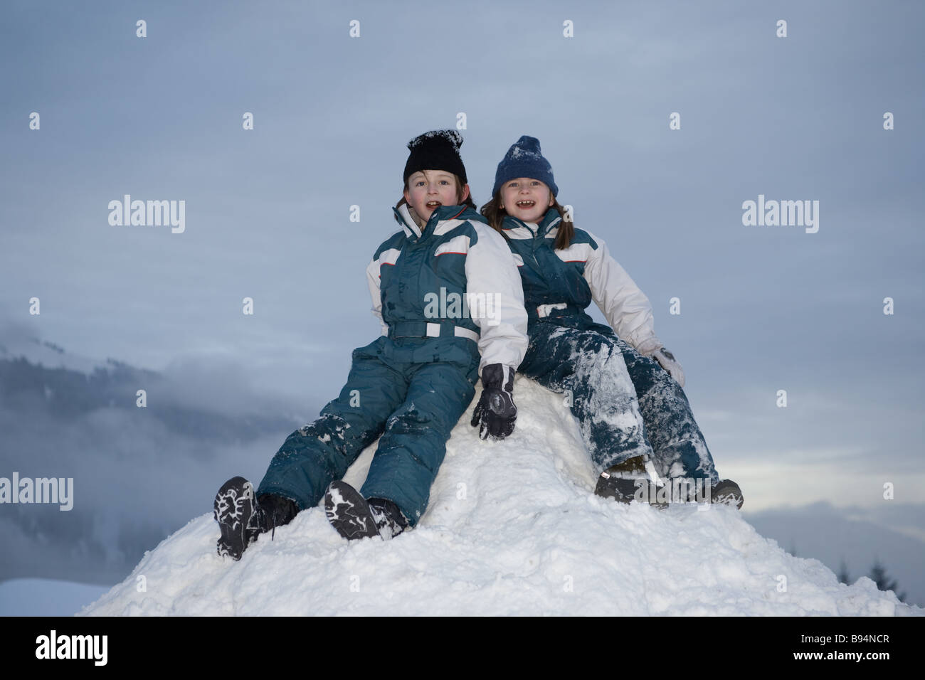 Two children, a boy and girl sit on a tall pile of snow with dark cloud ...
