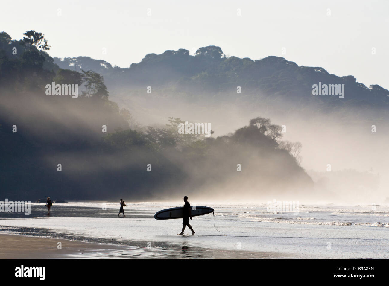 Surfers on the beach with sunlight shining through the morning mist at ...