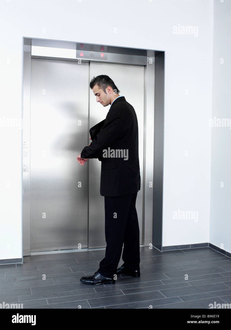 young businessman standing in front of elevator looking at his watch ...
