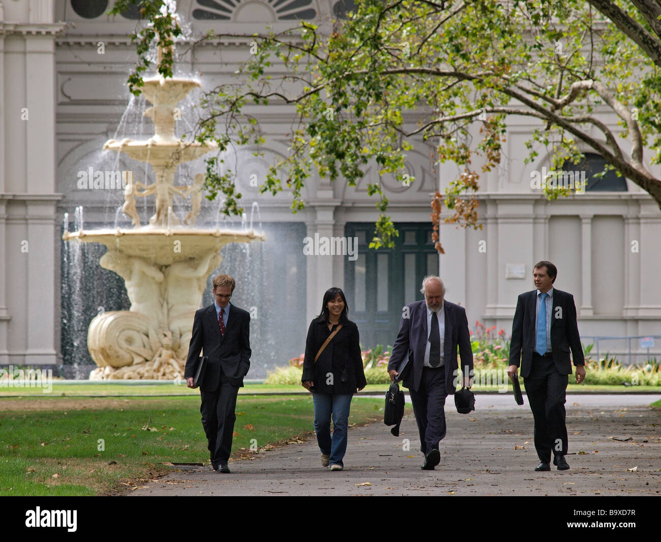 FOUR ADULTS IN LINE ABREAST WALKING THROUGH PARK WITH FOUNTAIN IN ...