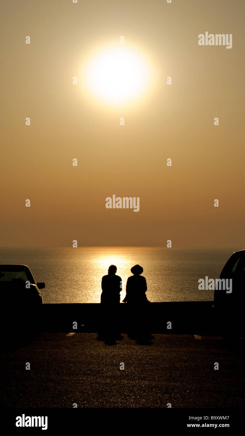 A silhouetted couple enjoy a sunrise over the sea from between parked ...