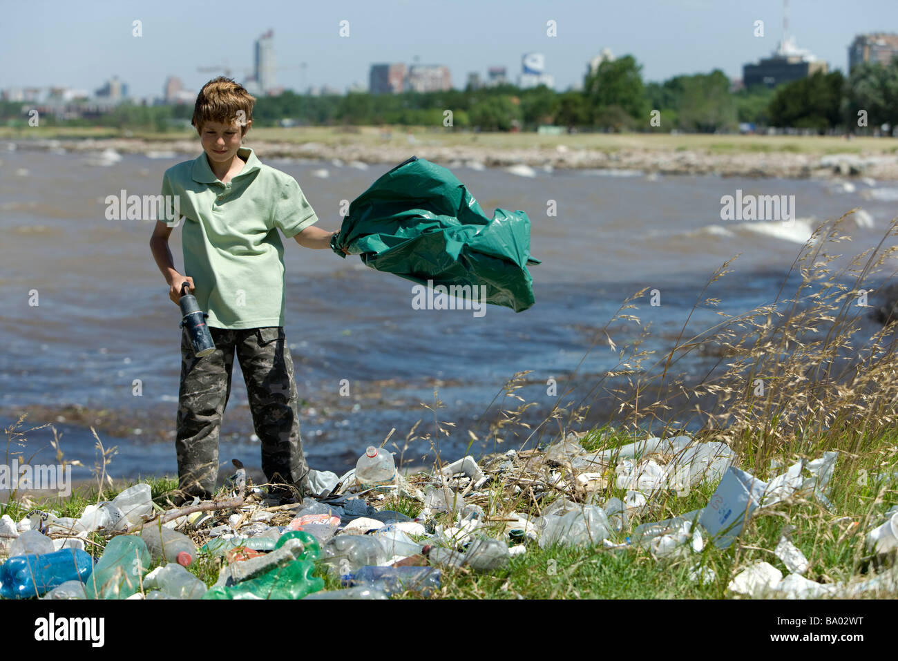 Boy picking up litter on shore, holding garbage bag and magnetic device ...