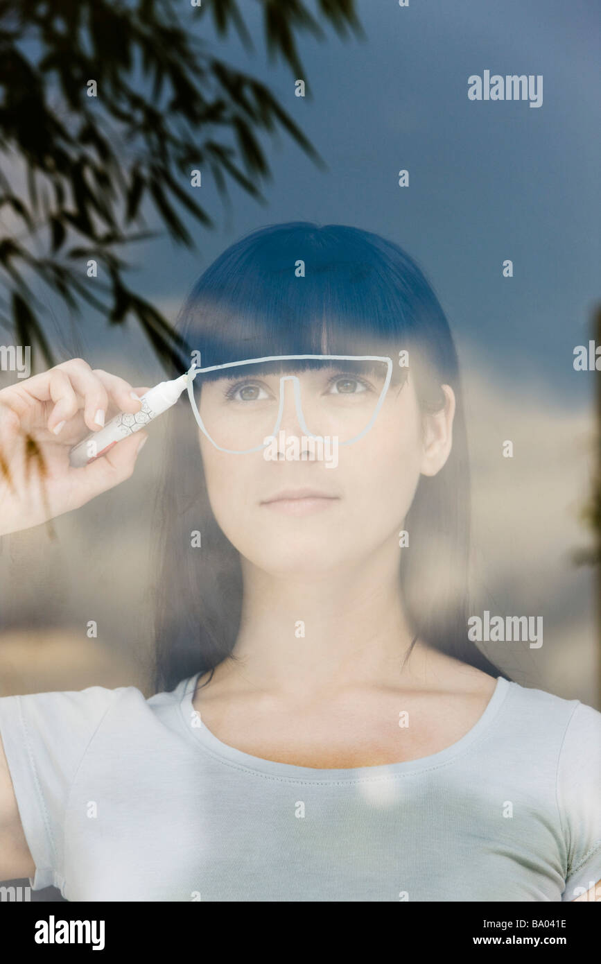 Young woman drawing a pair of glasses on a window pane, looking through ...