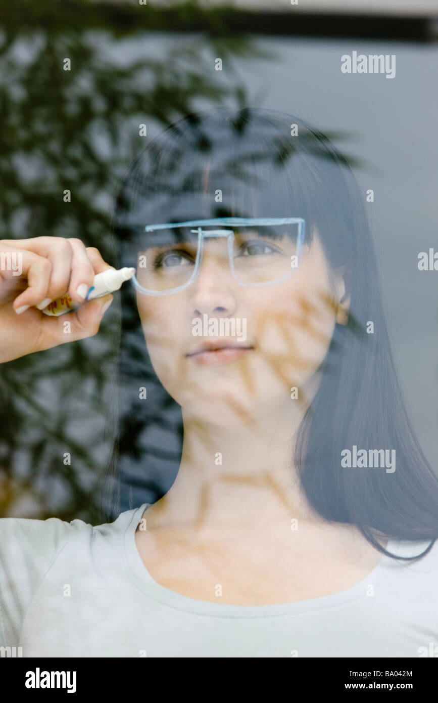 Young woman drawing a pair of glasses on a window pane, looking through ...