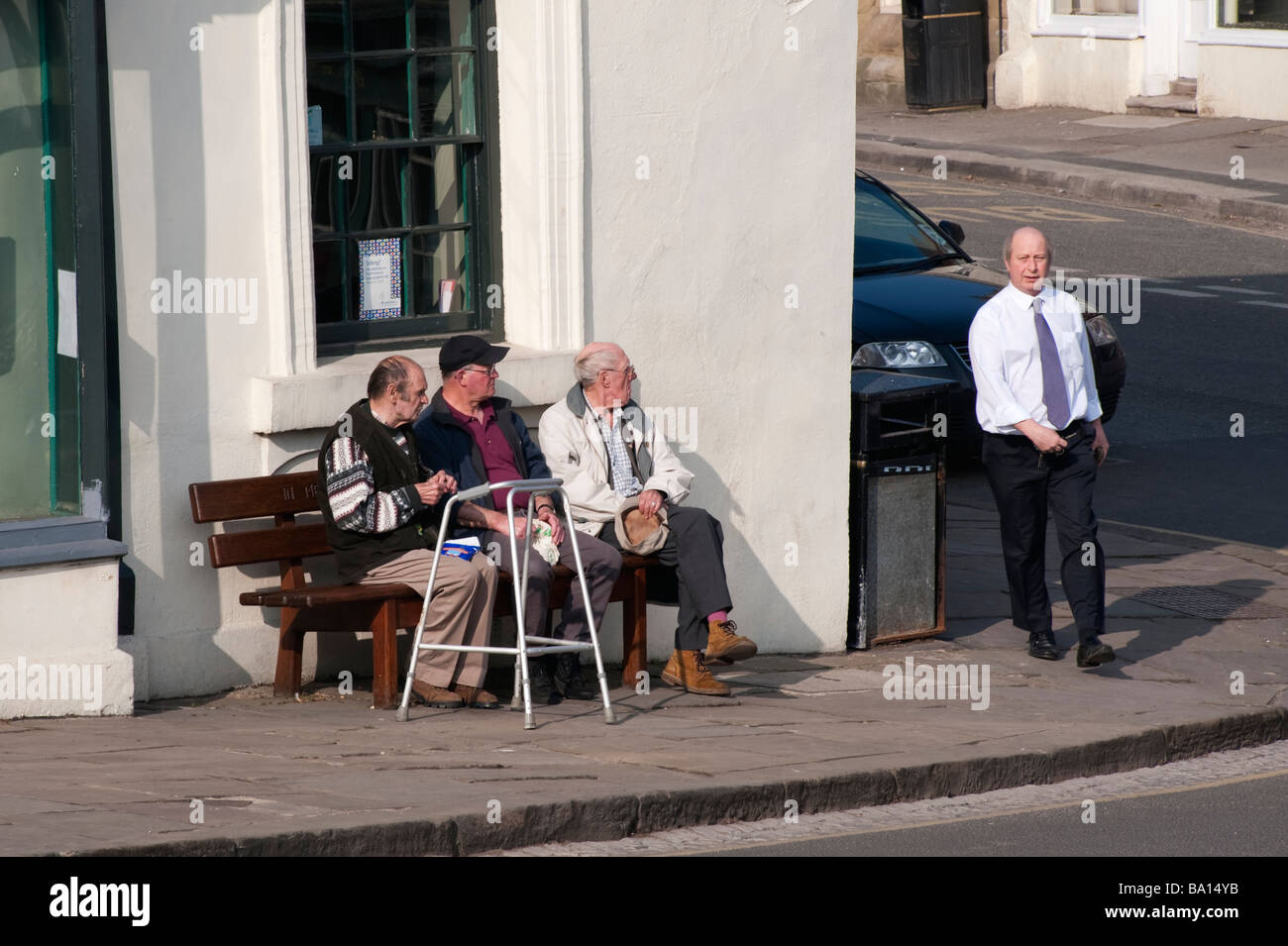 Three pensioners sitting on a street corner bench observing a passing ...