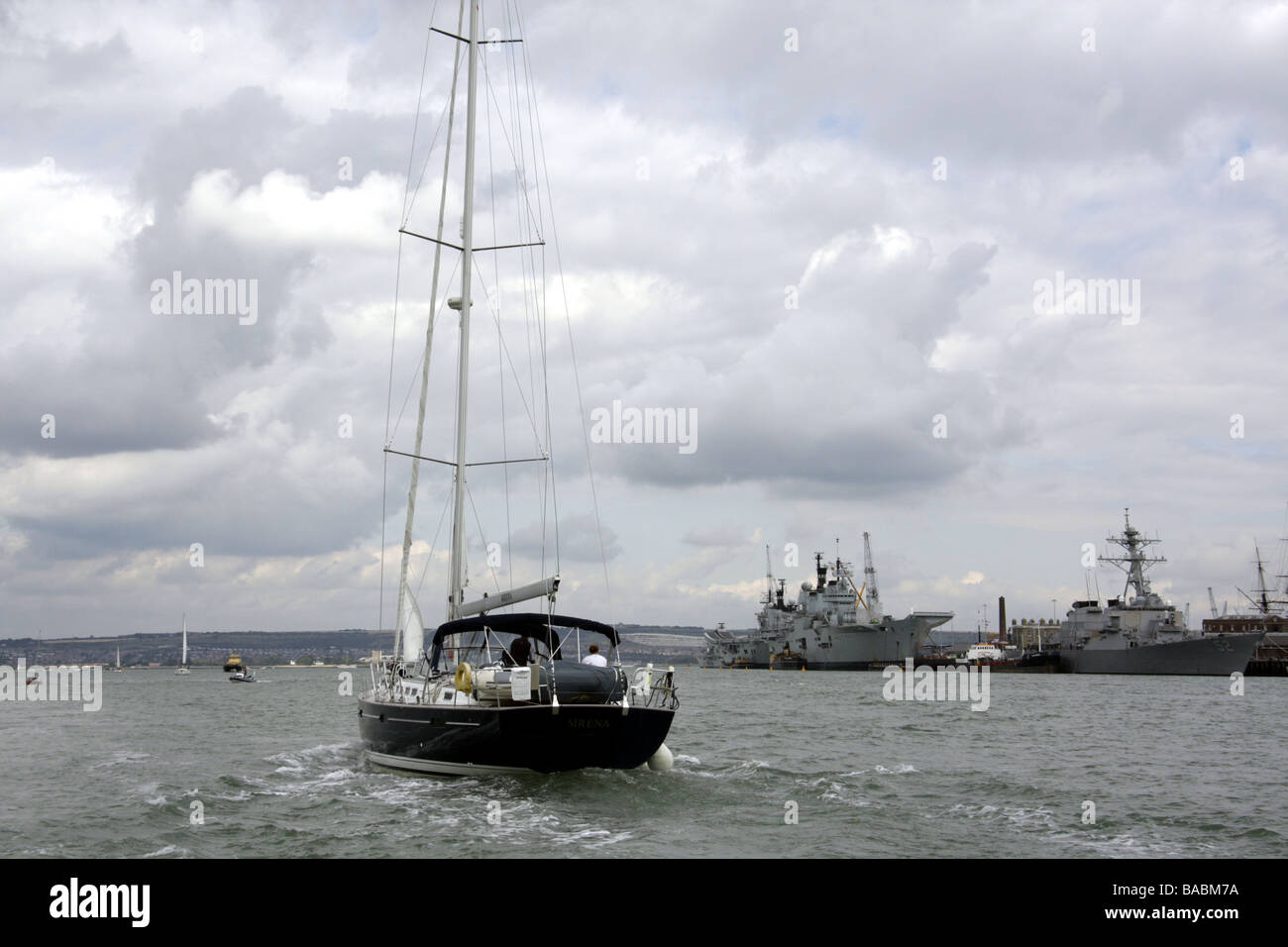 A small yacht under power in Portsmouth harbour with Royal Navy ships ...